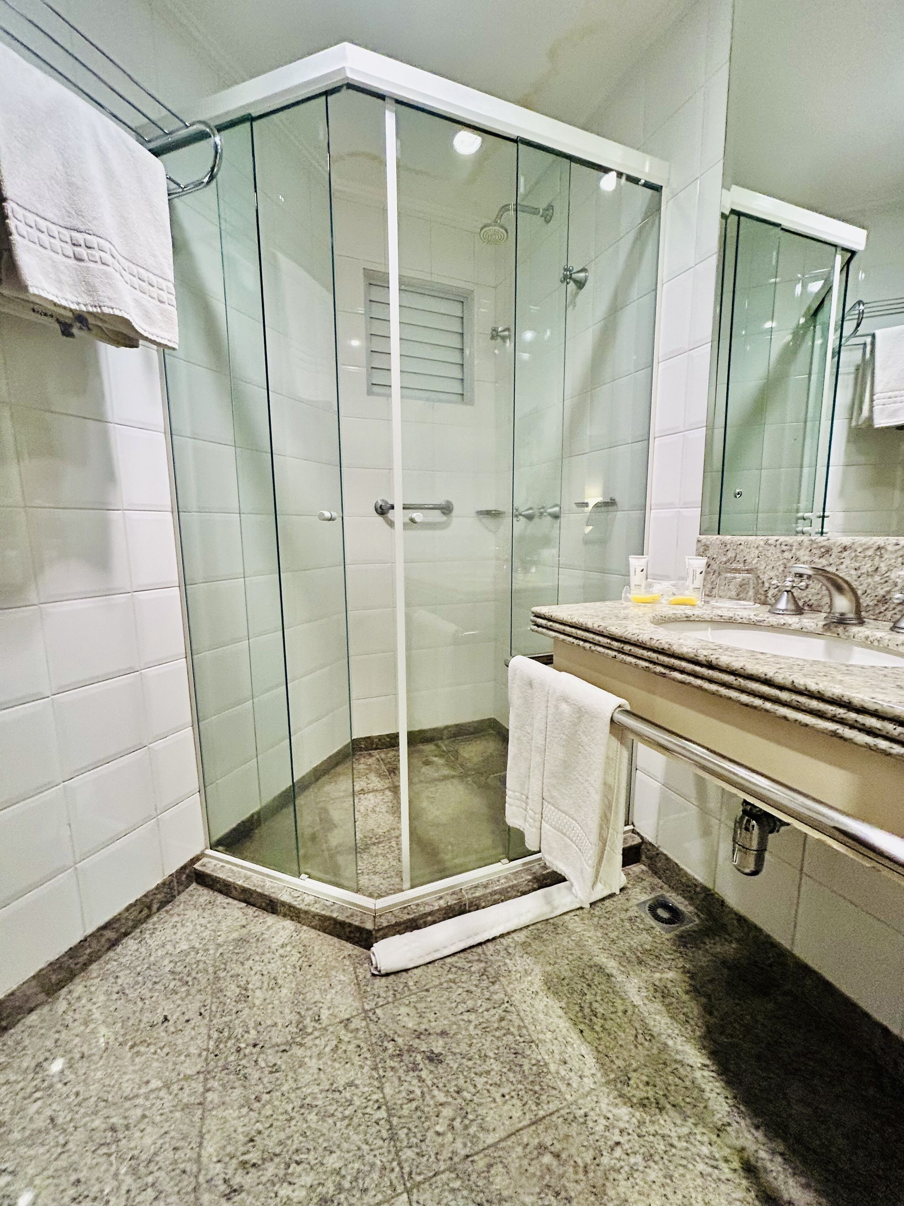Divisória de chuveiro envidraçada com uma toalha branca no chão, pia com bancada de granito e espelho acima da pia.