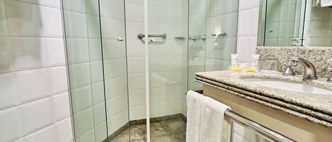 Divisória de chuveiro envidraçada com uma toalha branca no chão, pia com bancada de granito e espelho acima da pia.