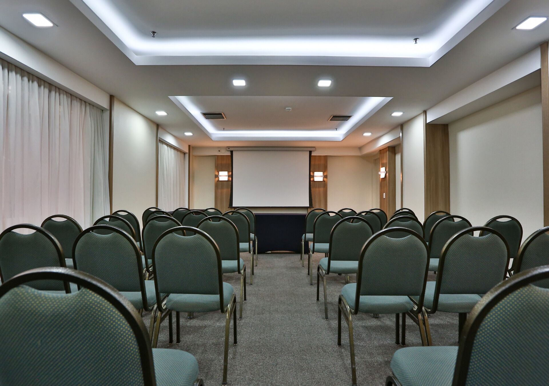 Uma sala de conferências com fileiras de cadeiras, uma tela de projetor e iluminação embutida no teto.
