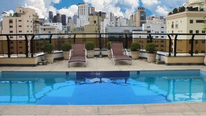 Piscina no terraço com duas espreguiçadeiras, plantas em vasos e vista para a cidade.