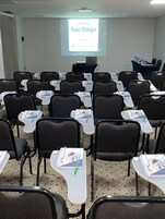 Uma sala de conferências com fileiras de cadeiras e mesas, uma tela de projetor exibindo 'San Diego' e uma mesa com garrafas de água e copos.