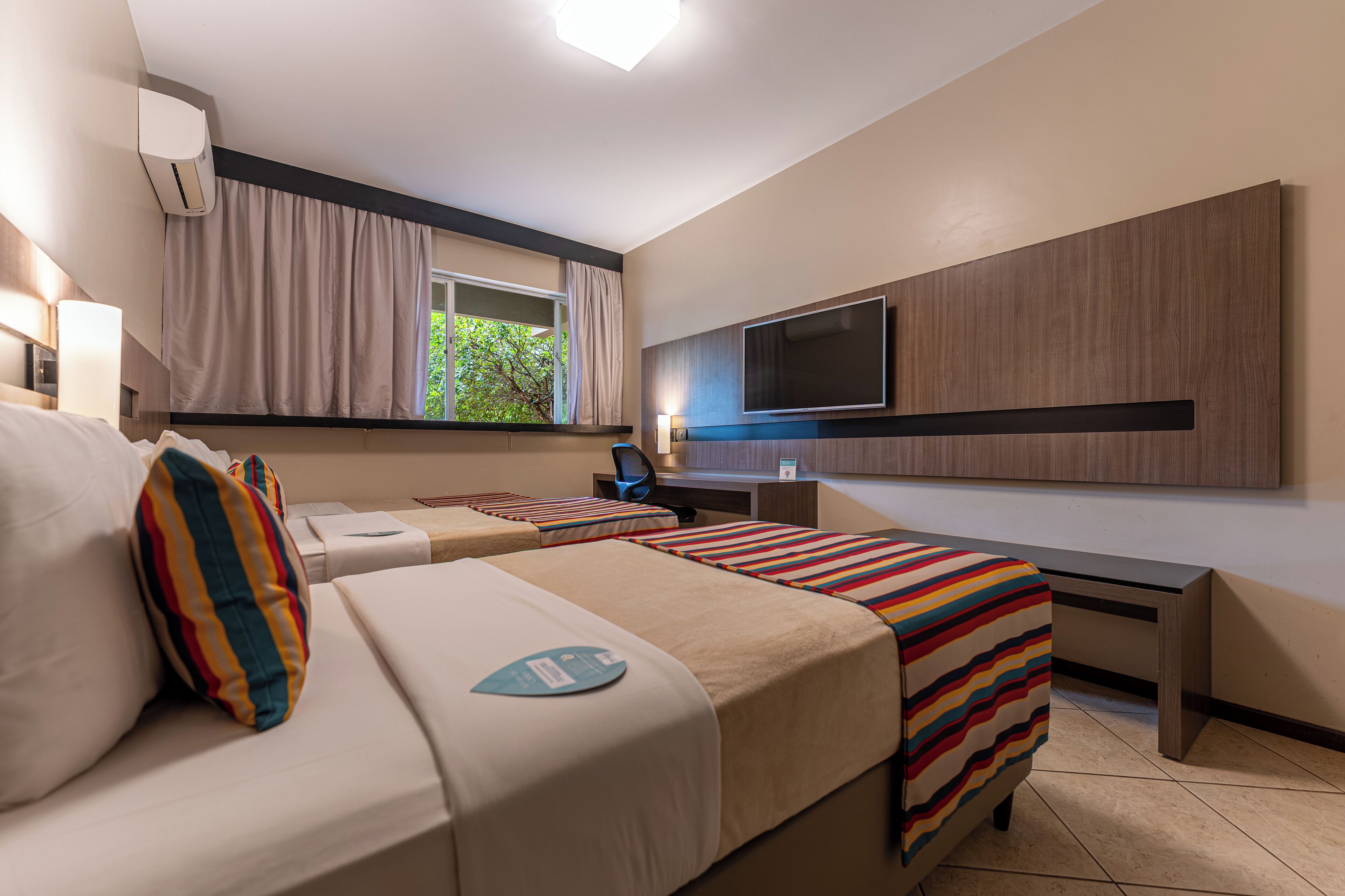Quarto de hotel com duas camas, uma escrivaninha, televisão e janela com cortinas.