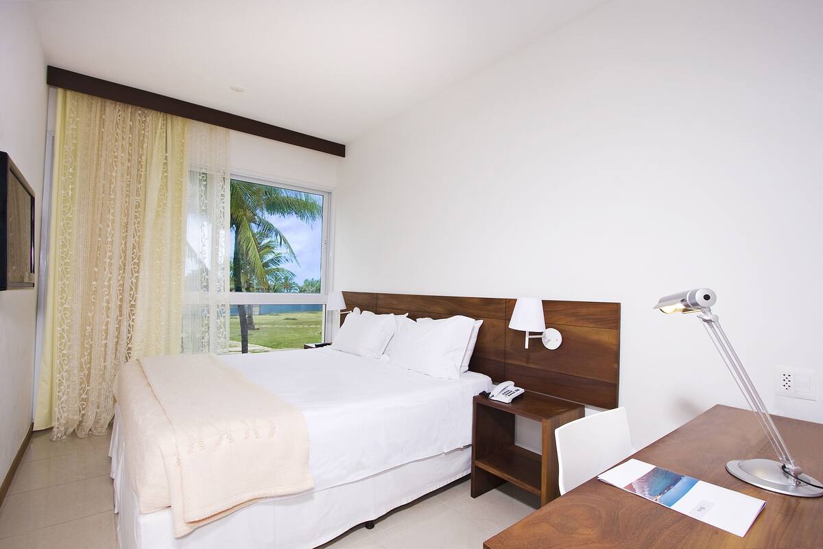 Quarto de hotel com uma cama grande, uma mesinha de cabeceira de madeira com um abajur, uma escrivaninha com uma cadeira e uma janela com cortinas.