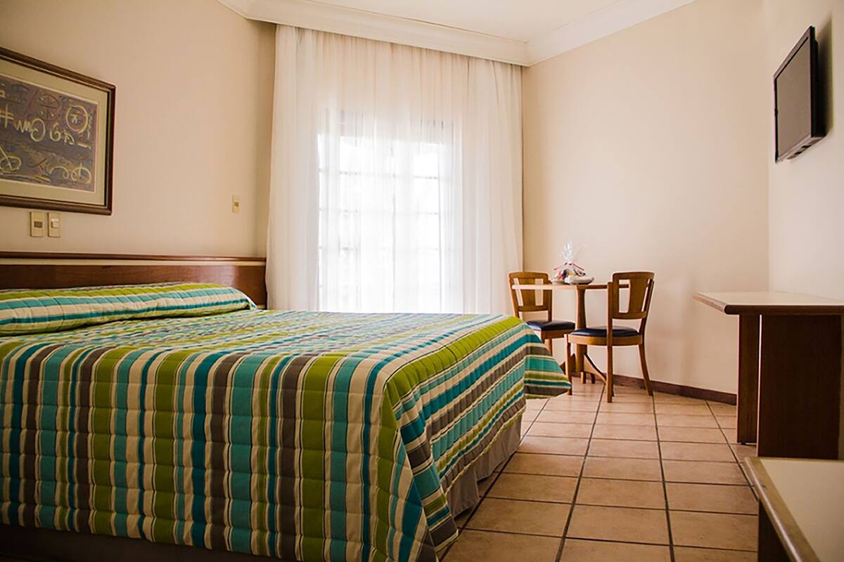 Quarto de hotel com cama, escrivaninha, duas cadeiras, televisão e janela com cortinas.