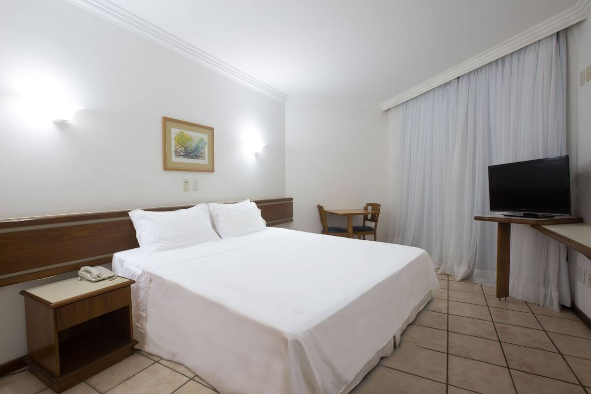 Quarto de hotel com uma cama grande, mesa de cabeceira com telefone, uma cadeira, escrivaninha com televisão e um quadro na parede.