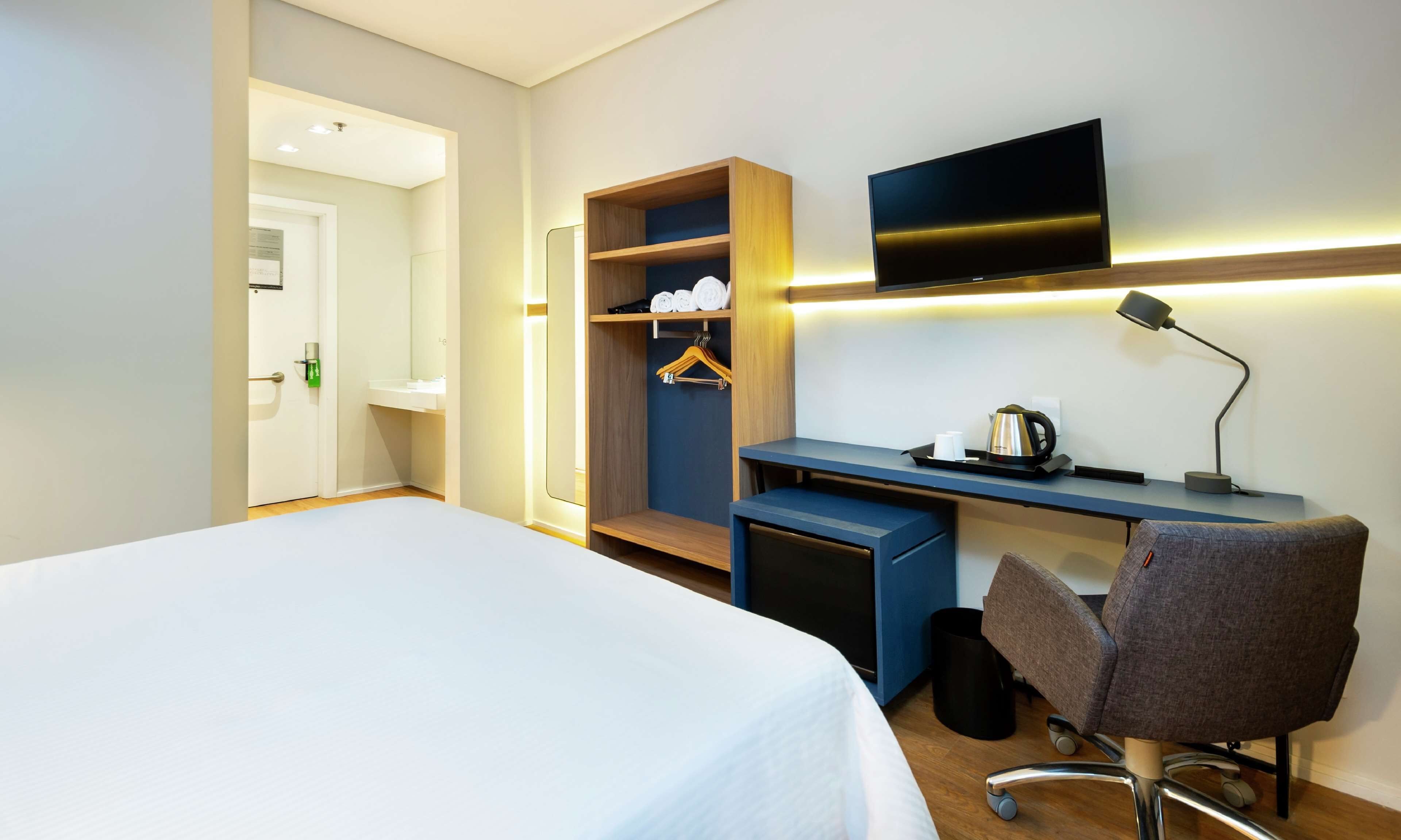 Quarto de hotel com uma cama, uma escrivaninha com cadeira, televisão, prateleira com roupas e um bule, além de uma pequena lixeira.