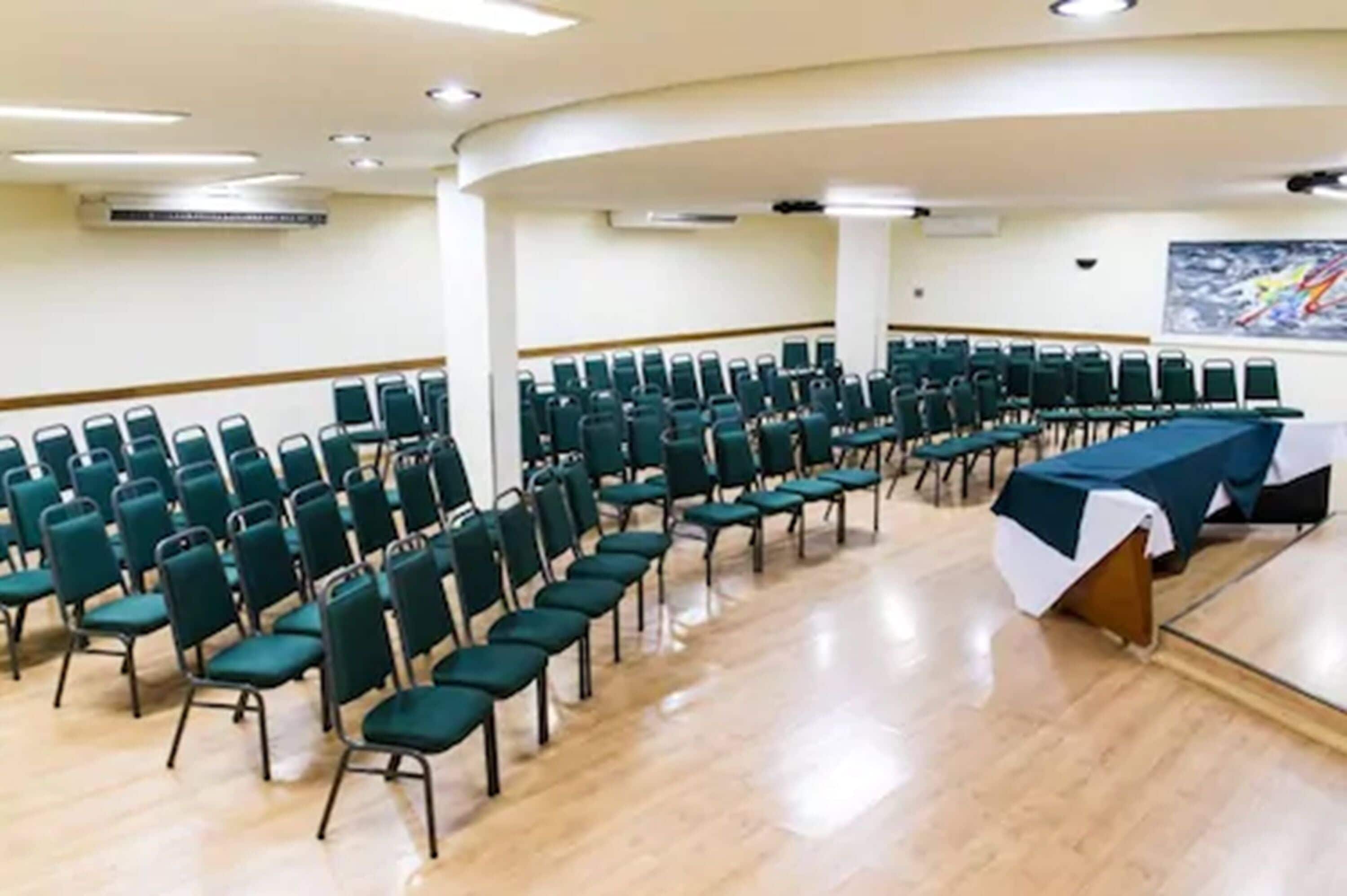 Uma sala de conferências com fileiras de cadeiras verdes e uma mesa comprida coberta com toalha de mesa branca e verde-água.