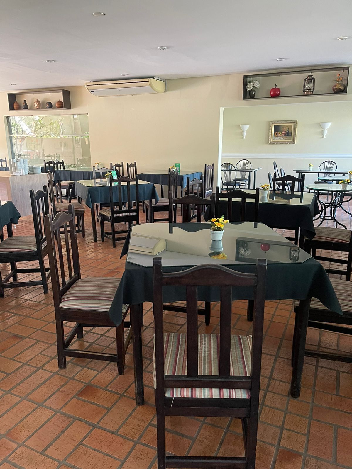 Uma área de refeições com várias mesas e cadeiras, algumas com toalhas. Há prateleiras com objetos decorativos nas paredes.