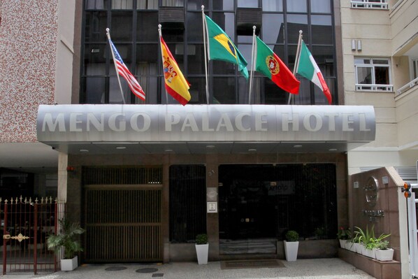 Entrada do Hotel Mengo Palace, com bandeiras, portão e plantas em vasos.