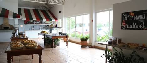 Um café com chão de azulejos, uma mesa de madeira com comida, um forno de tijolos e um letreiro com ilustração de frutas.