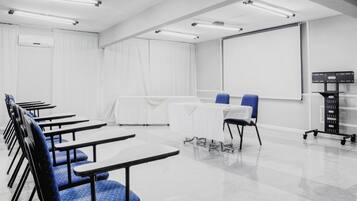 Uma sala de conferência com fileiras de cadeiras azuis, uma mesa branca e uma tela de projeção.