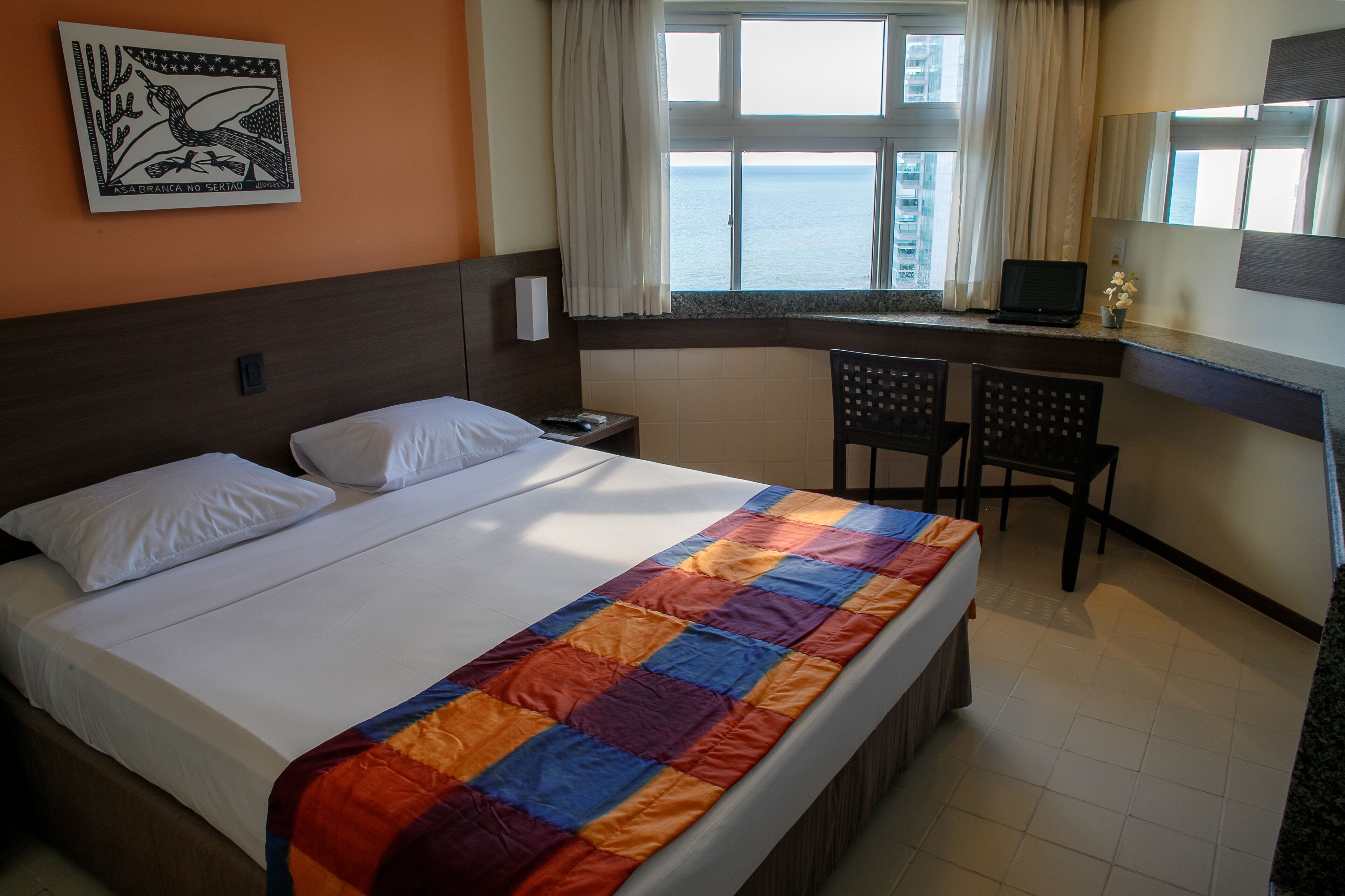 Quarto de hotel com cama, escrivaninha com laptop, cadeira e janela com cortinas.
