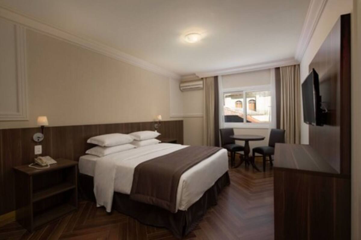 Quarto de hotel com cama, mesa de cabeceira, televisão e um pequeno espaço de refeições com mesa e cadeiras.