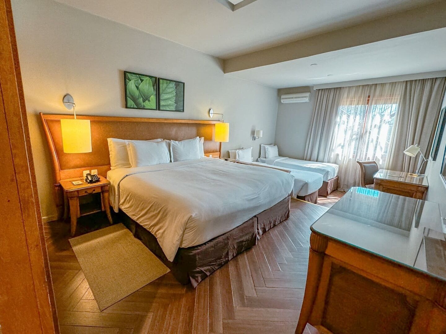 Quarto de hotel com uma cama grande, duas mesas de cabeceira, uma escrivaninha e uma cadeira. Há duas luminárias na parede, uma janela com cortinas e um aparelho de ar condicionado.