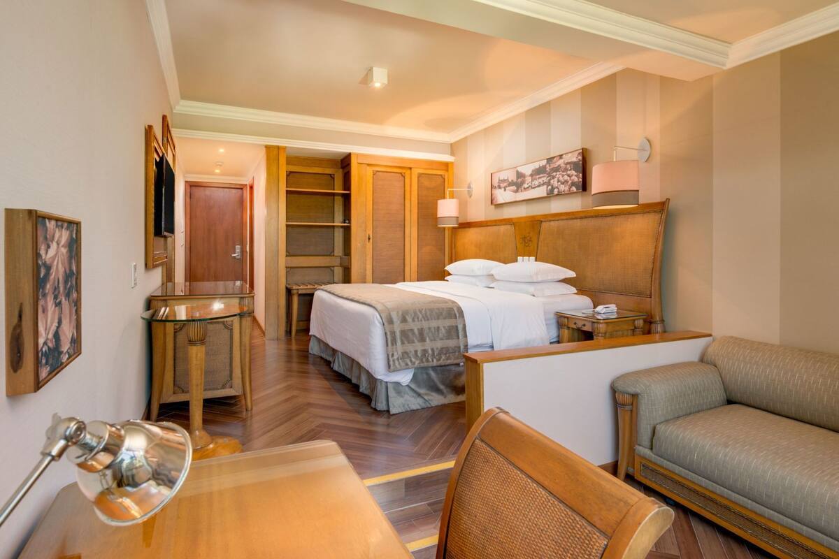 Quarto de hotel com cama, escrivaninha, poltrona, sofá, espelho e guarda-roupa.