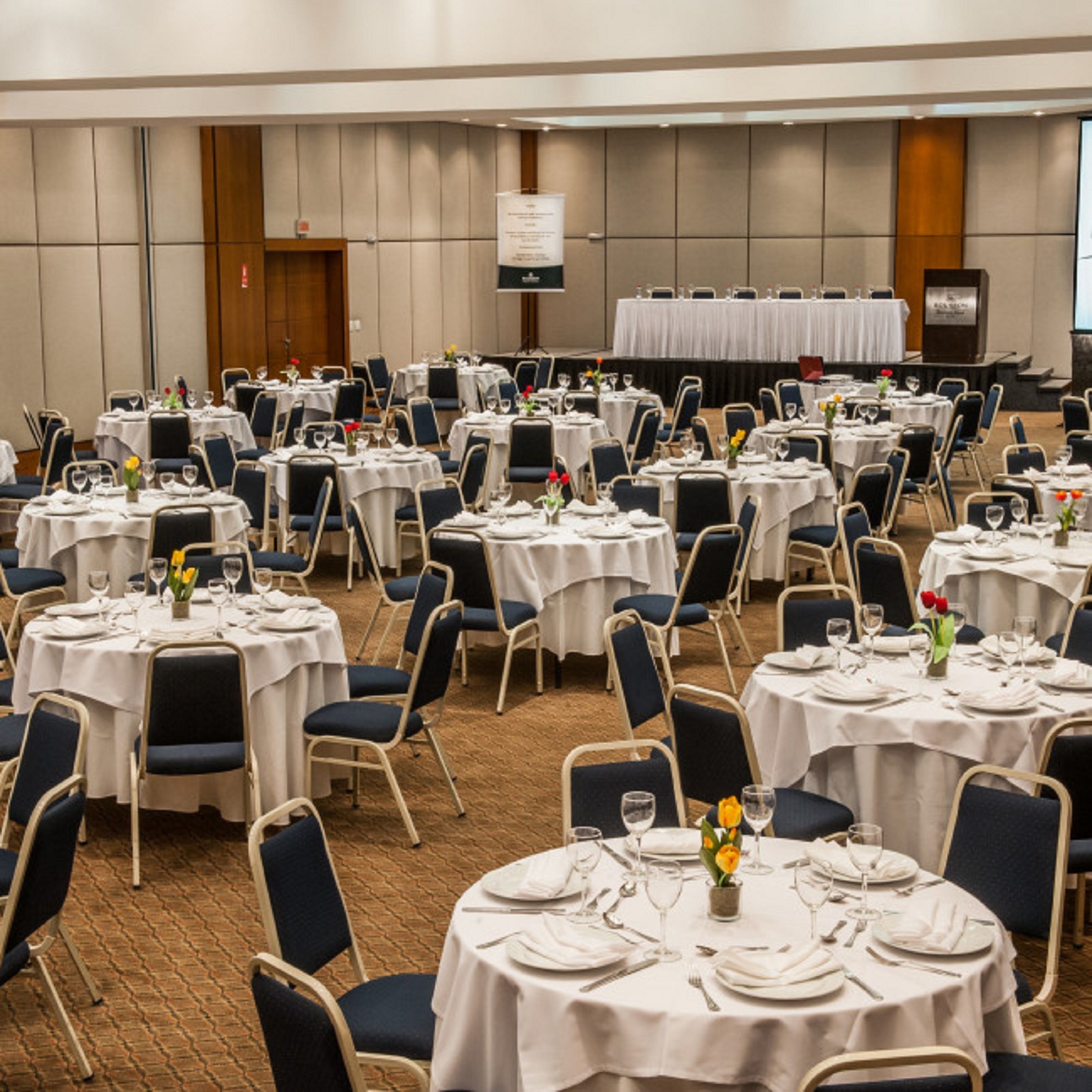 Uma sala de banquetes com mesas redondas dispostas para refeições, cadeiras e um palco com uma tela.