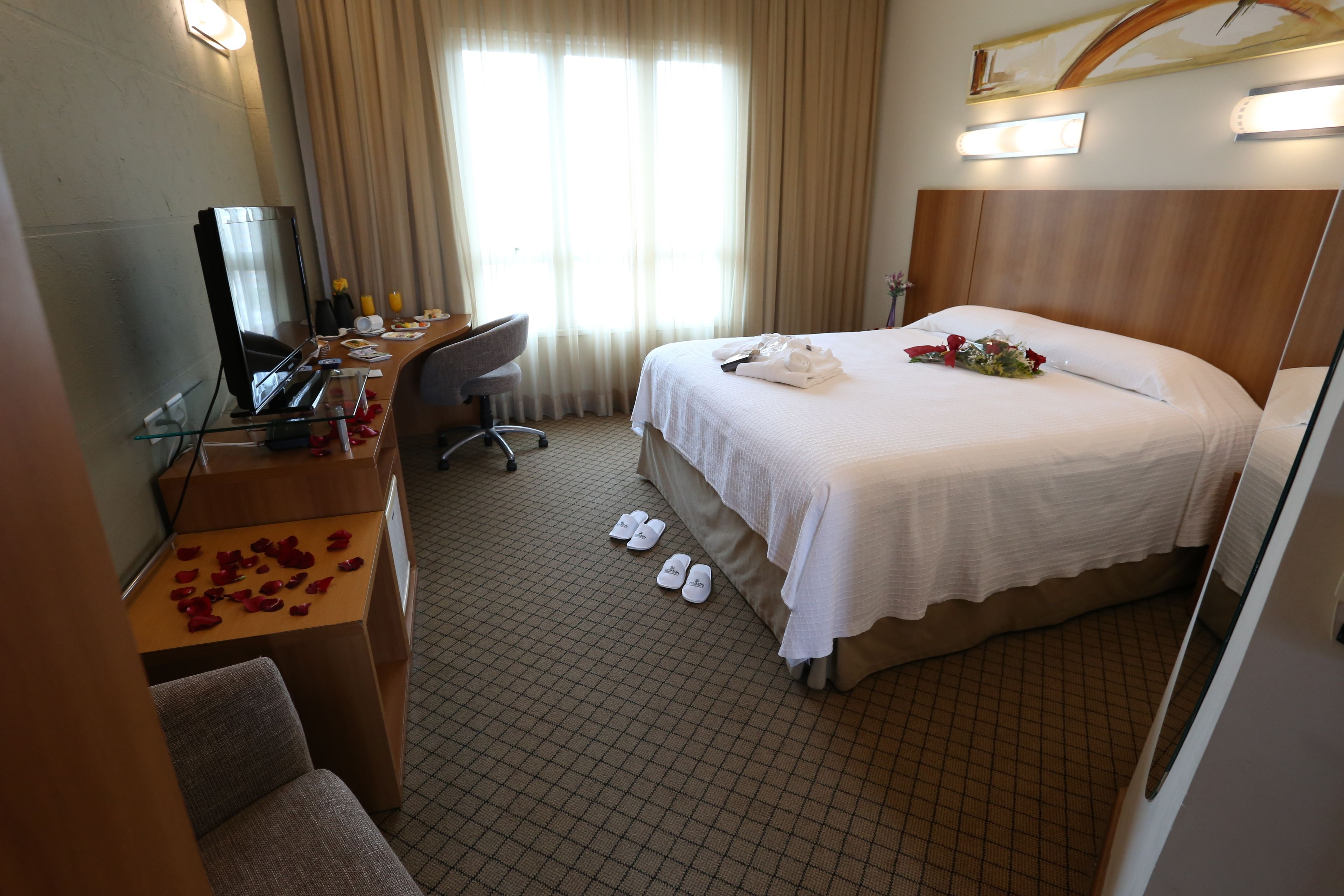 Quarto de hotel com cama, escrivaninha com computador, cadeira e janela com cortinas.
