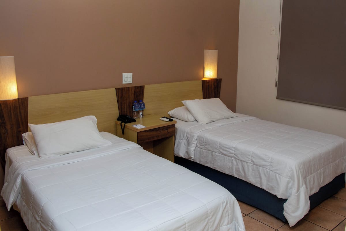 Duas camas de solteiro com roupa de cama branca, uma cabeceira de madeira, mesas de cabeceira com luminárias e uma TV fixada na parede.