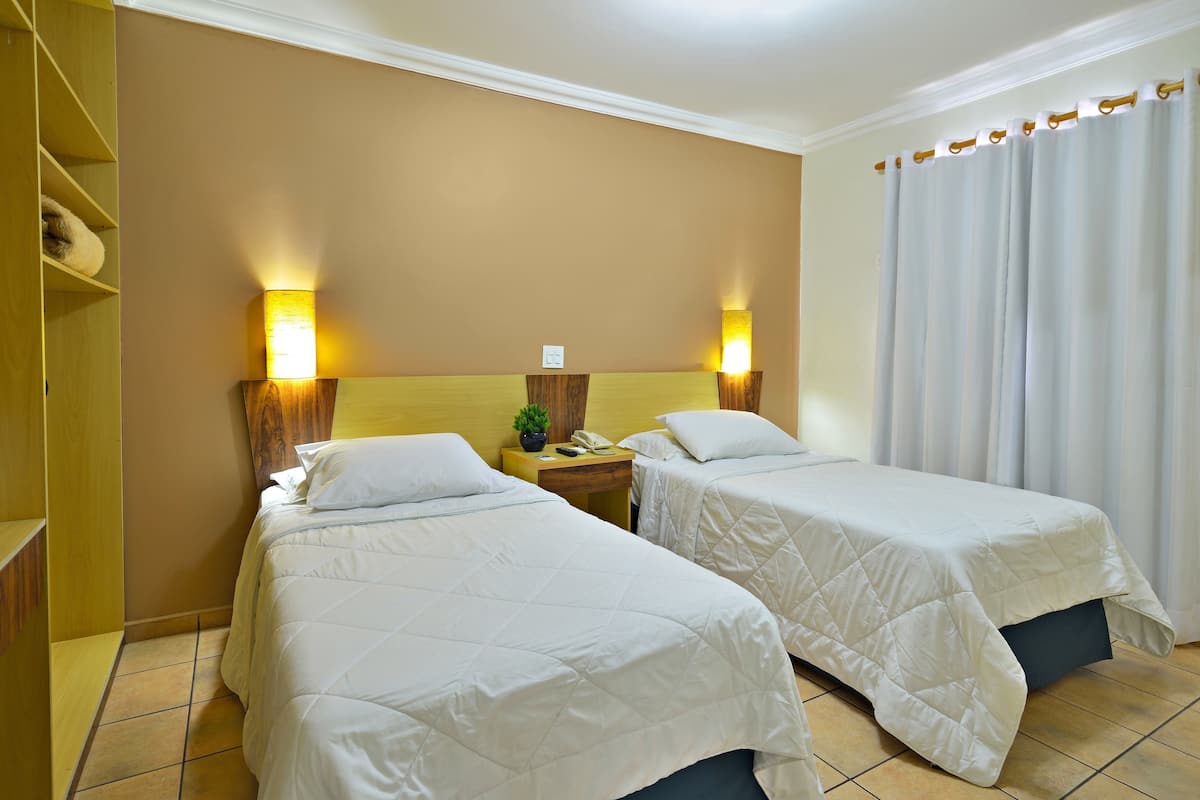 Quarto de hotel com duas camas, um guarda-roupa de madeira, uma mesa de cabeceira com abajur e uma janela com cortinas.