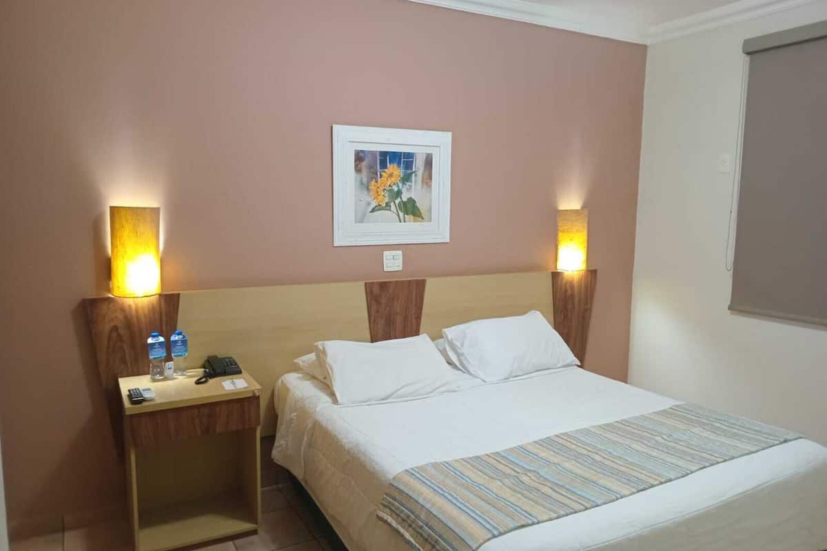Quarto de hotel com uma cama, mesas de cabeceira, um quadro e luminárias na parede.