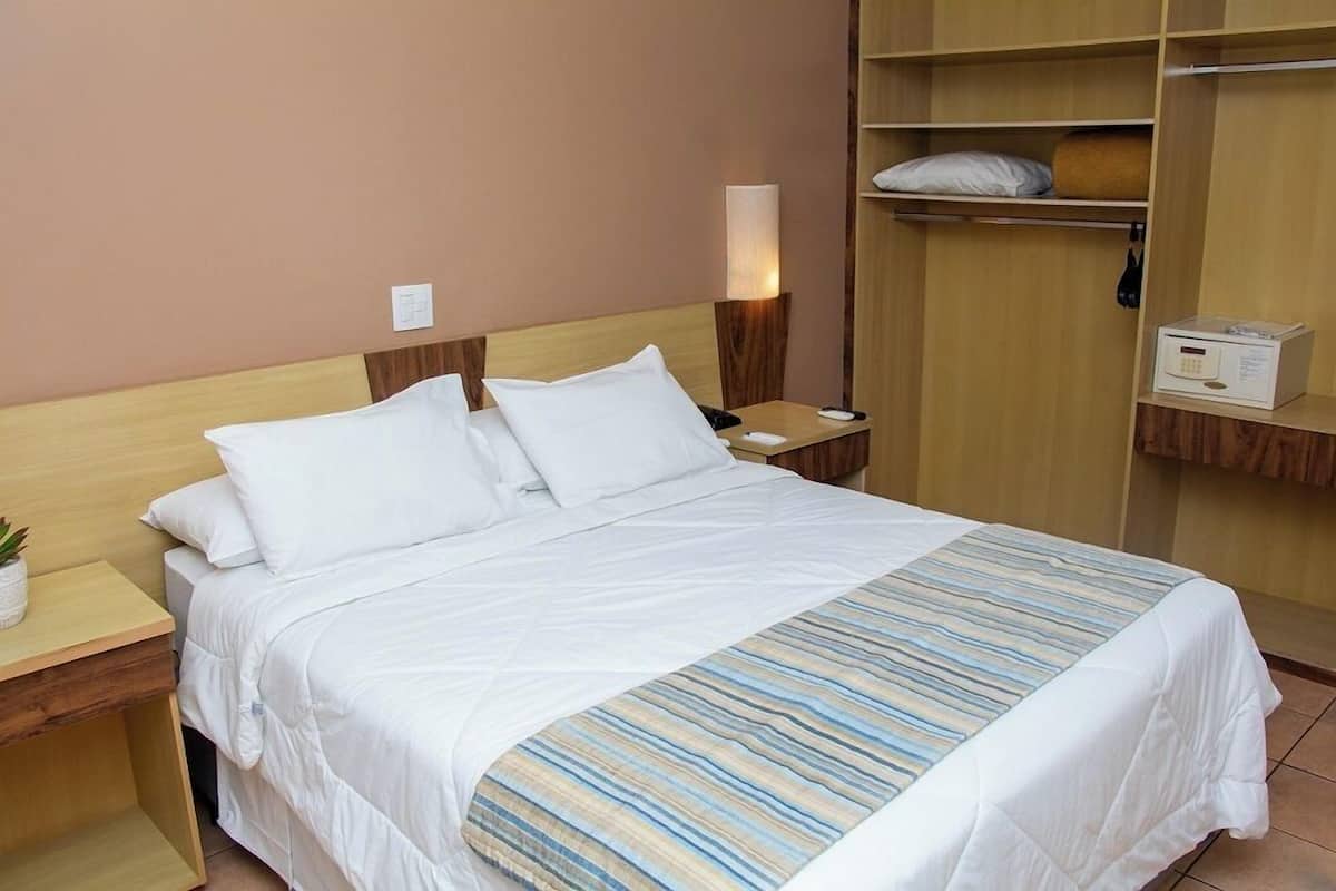 Quarto de hotel com cama, mesas de cabeceira, abajur, guarda-roupa e um pequeno objeto fixado na parede.