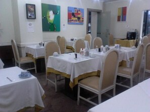 Uma área de refeições com várias mesas e cadeiras, toalhas de mesa brancas e algumas pinturas nas paredes.
