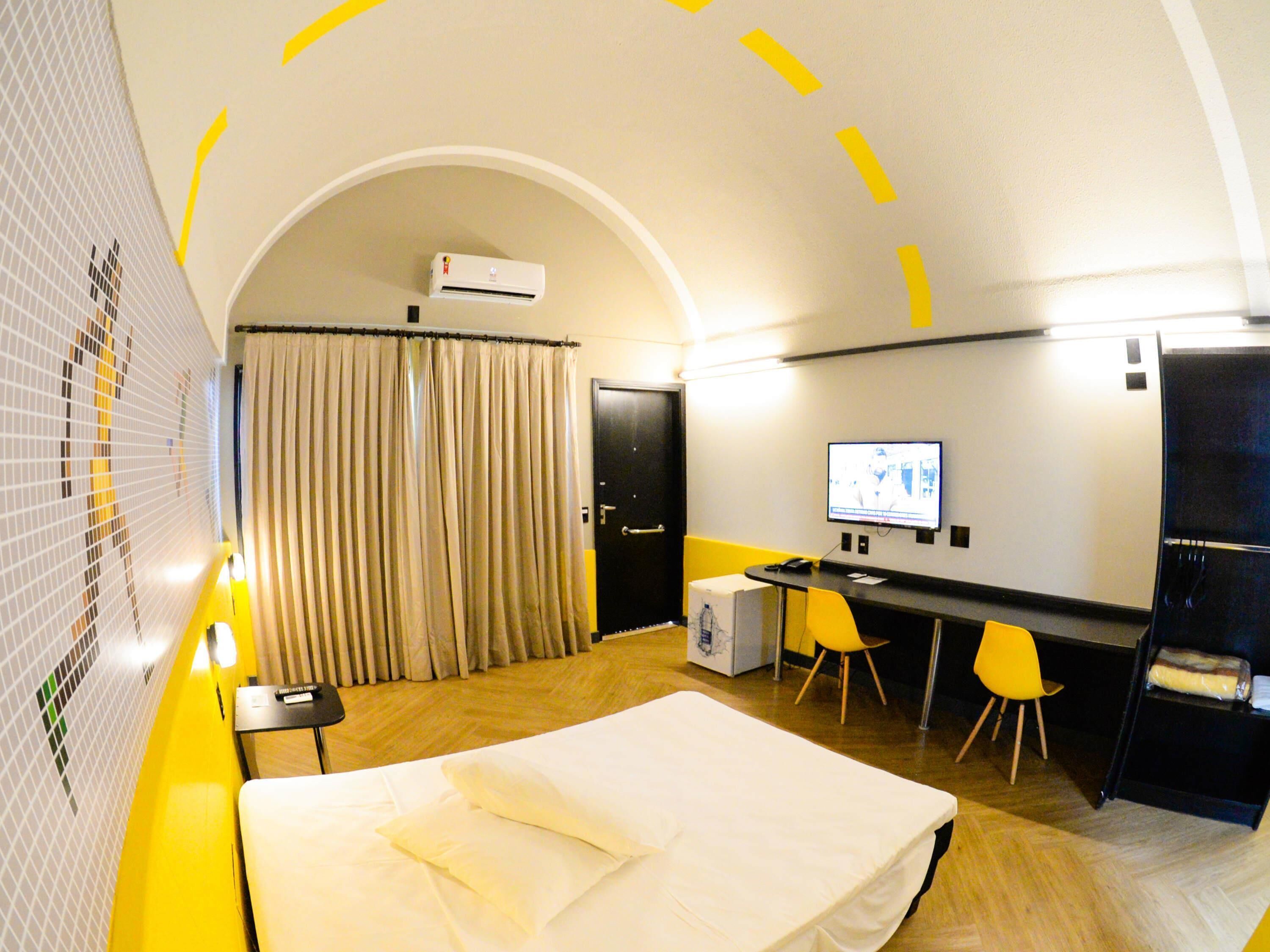Quarto de hotel com cama, escrivaninha, cadeira, aparelho de ar condicionado e televisão.