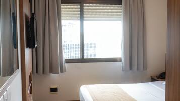 Quarto de hotel com cama, escrivaninha, cadeira, televisão, janela com cortinas e ar condicionado.