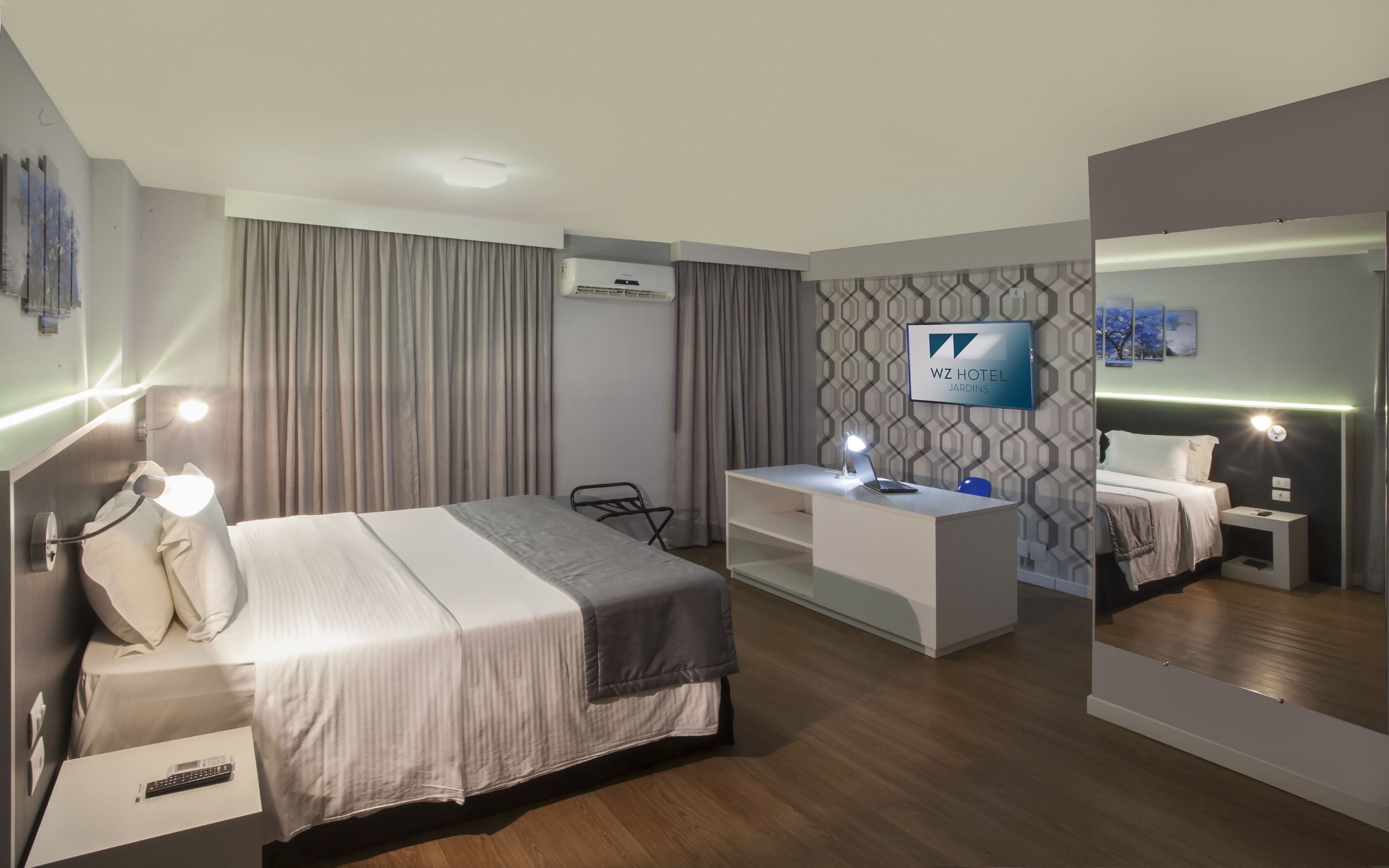Quarto de hotel com cama, mesa de cabeceira, escrivaninha e espelho.