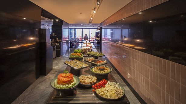 Disposição de buffet em um corredor moderno de hotel, com diversos alimentos e vista para a área de refeições ao fundo.