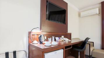 Quarto de hotel com escrivaninha de madeira, uma cadeira, um pequeno frigorífico, máquina de café e televisão na parede.