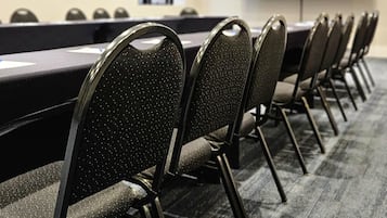 Uma sala de conferências com uma mesa comprida, fileiras de cadeiras, uma tela de projetor e uma janela.