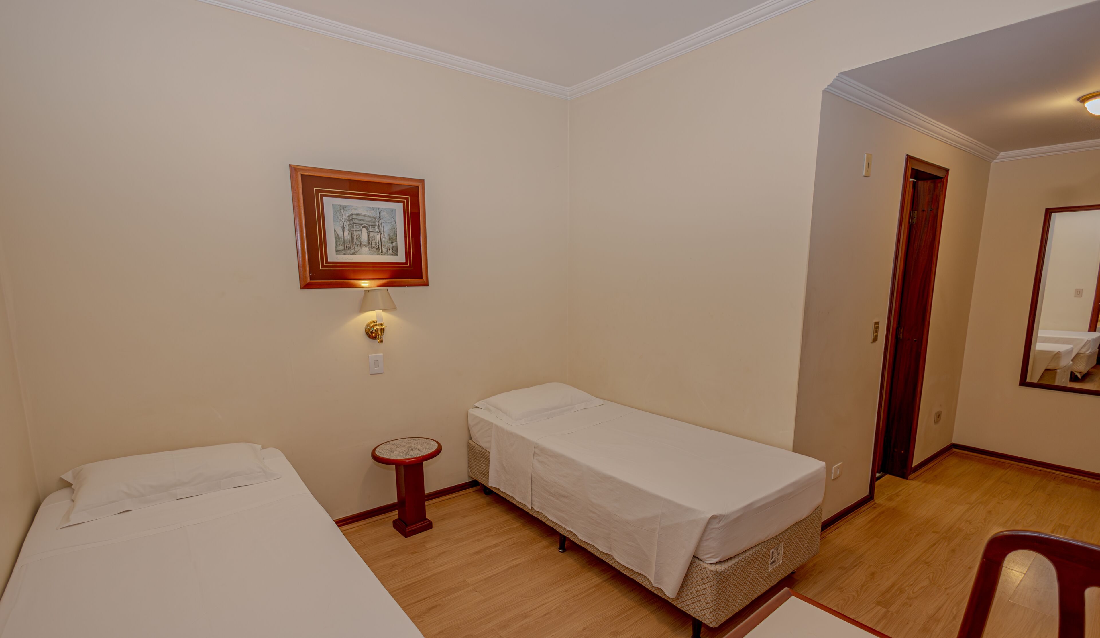 Quarto pequeno e limpo com duas camas de solteiro, uma mesa redonda pequena, um abajur fixado na parede e um quadro emoldurado.