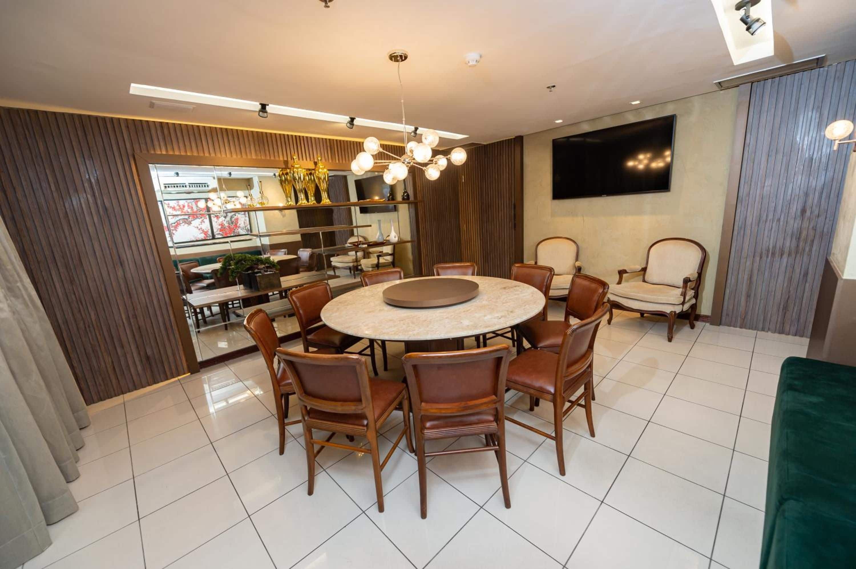 Uma sala de jantar com uma mesa redonda cercada por cadeiras, um candelabro e uma televisão fixada na parede.