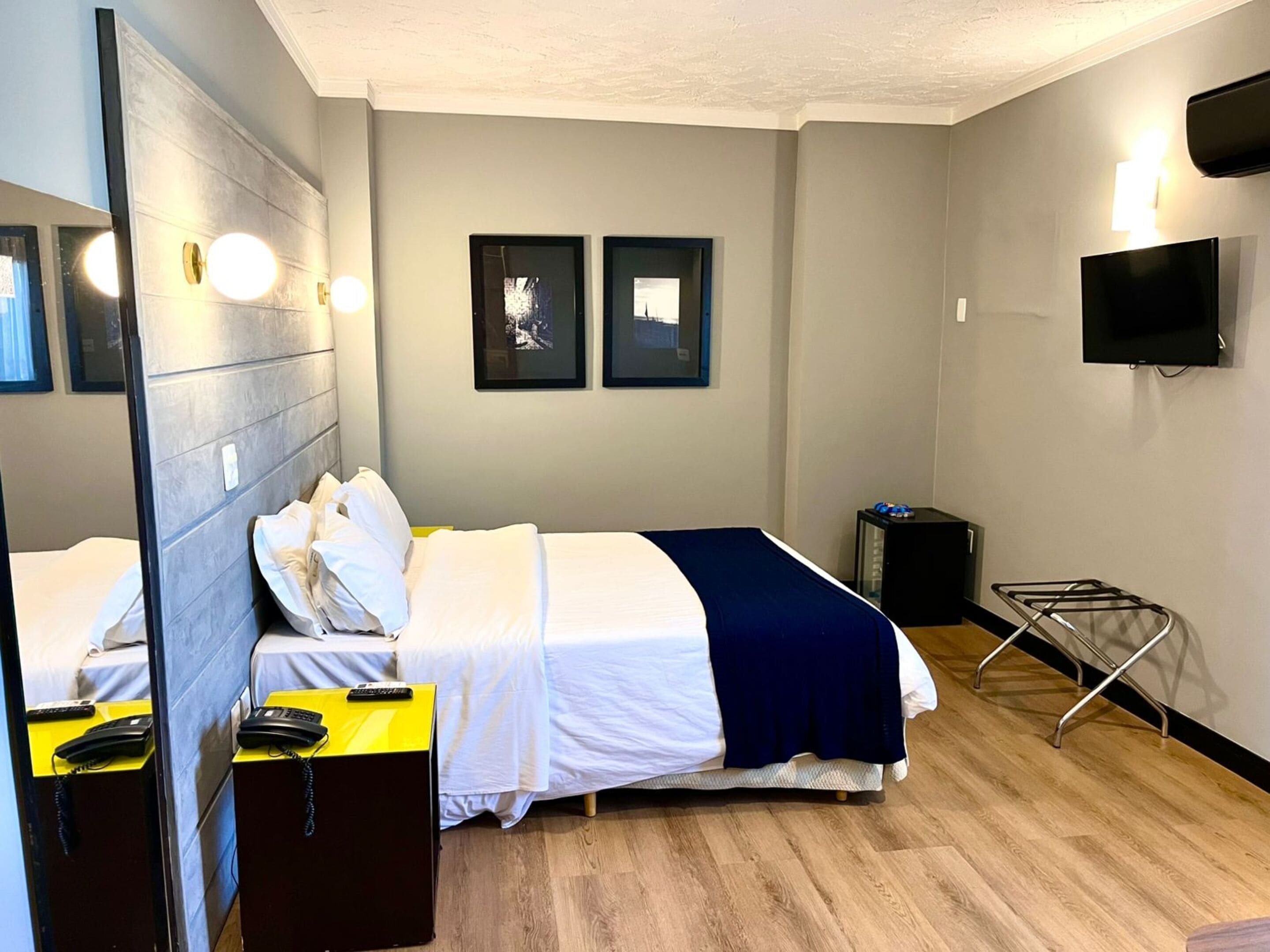 Quarto de hotel com cama, mesas de cabeceira, espelho, televisão de tela plana e uma pequena mesa com um alto-falante.