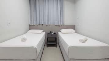 Quarto pequeno e limpo, com duas camas de solteiro, uma mesinha com telefone e janela com cortinas.