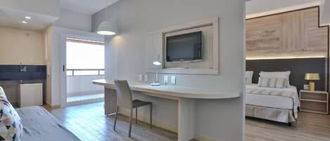 Quarto de hotel moderno com cama, escrivaninha com televisão, cadeira e mesa pequena.
