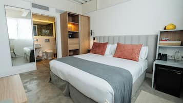 Quarto de hotel moderno com uma cama grande, guarda-roupa, secretária pequena e mini-frigorífico.