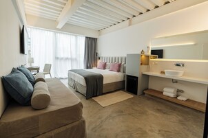 Quarto de hotel moderno com cama, sofá, mesa de cabeceira, luminária, espelho e área de banheiro com pia e porta-toalhas.
