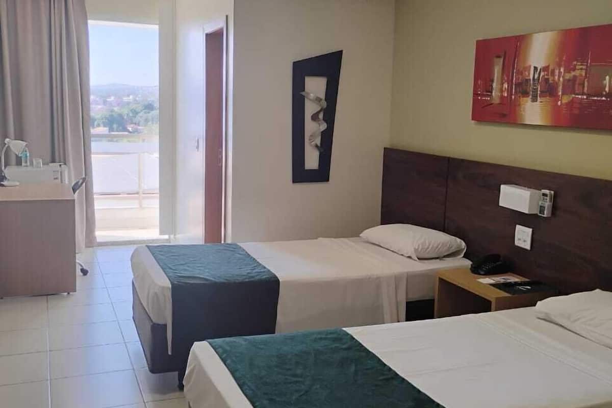 Quarto de hotel com duas camas, uma escrivaninha, ar condicionado e um quadro na parede.