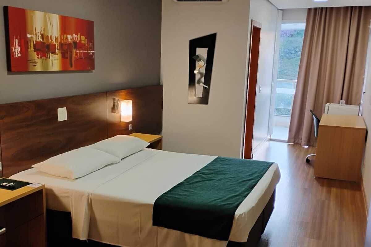 Quarto de hotel com piso de madeira, cama com roupa de cama branca e manta verde, mesa de cabeceira com luminária, quadro na parede, aparelho de ar condicionado e vista para o exterior através de uma janela com cortinas.