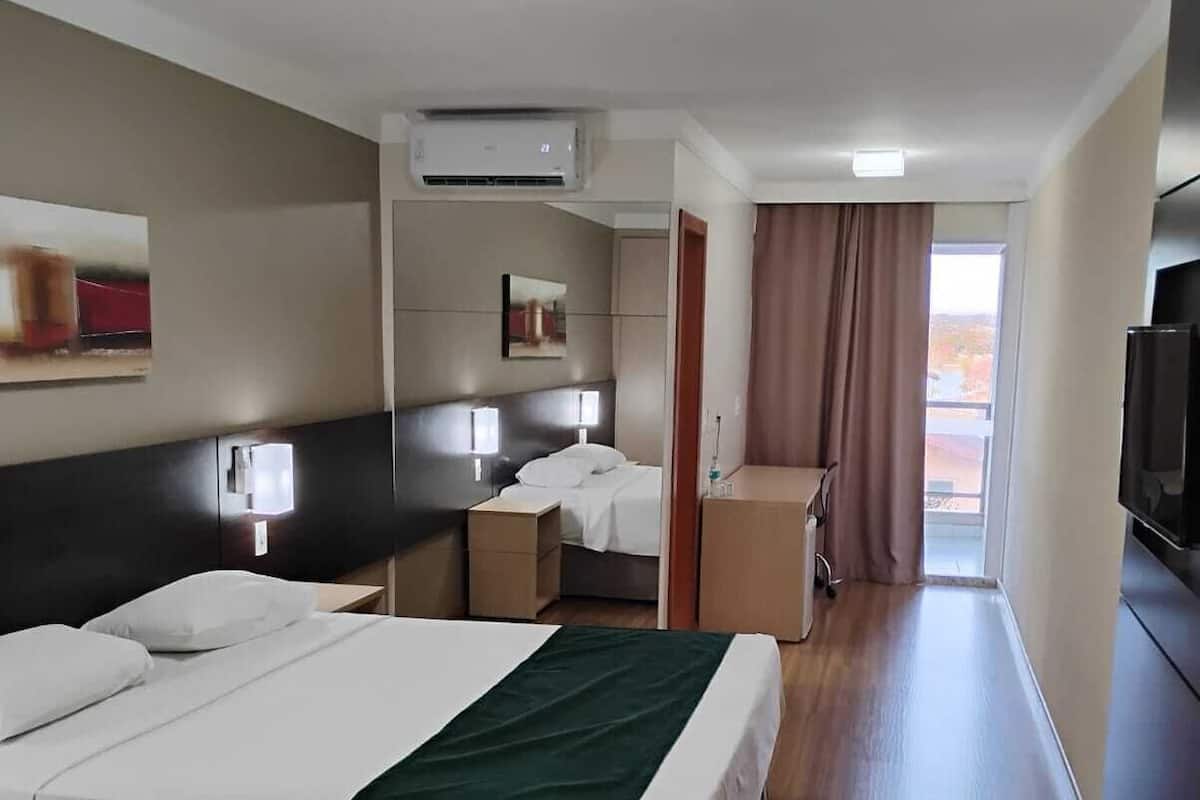 Quarto de hotel com cama, mesas de cabeceira, escrivaninha, televisão e ar condicionado.