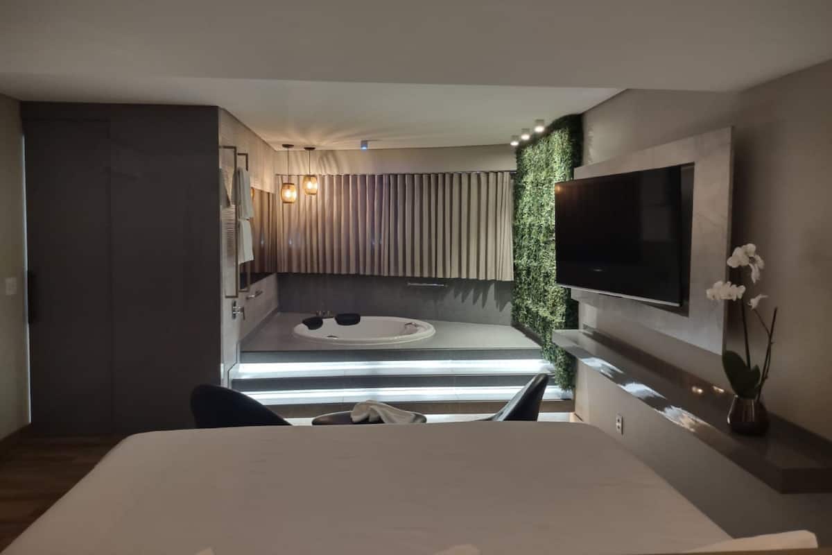 Quarto de hotel moderno com uma cama grande, uma TV fixada na parede, um pequeno espaço de estar com mesa e cadeiras, e vista para a cidade pela janela.