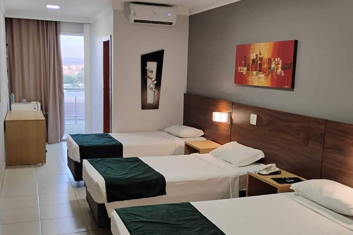 Quarto de hotel com três camas de solteiro, cabeceira de madeira, um quadro na parede e vista da cidade pela janela.