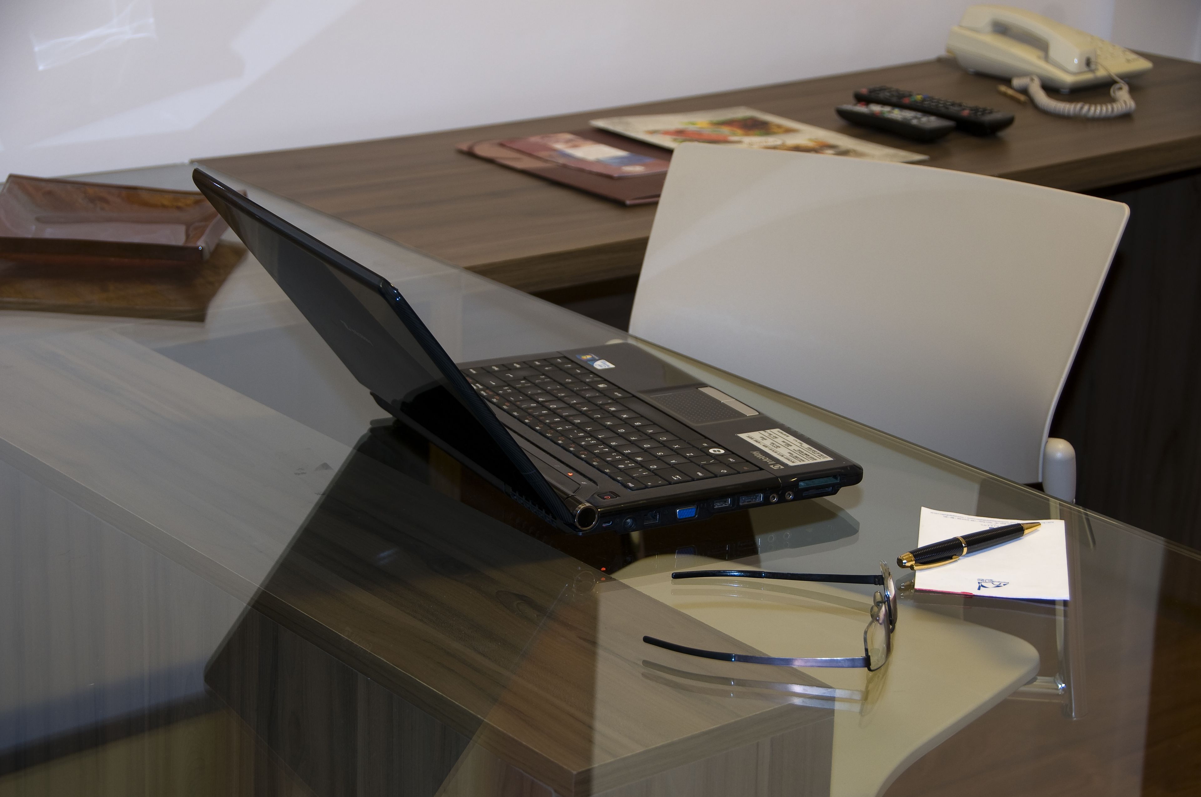 Um laptop sobre uma mesa de vidro, com uma caneta, óculos e um bloco de notas.
