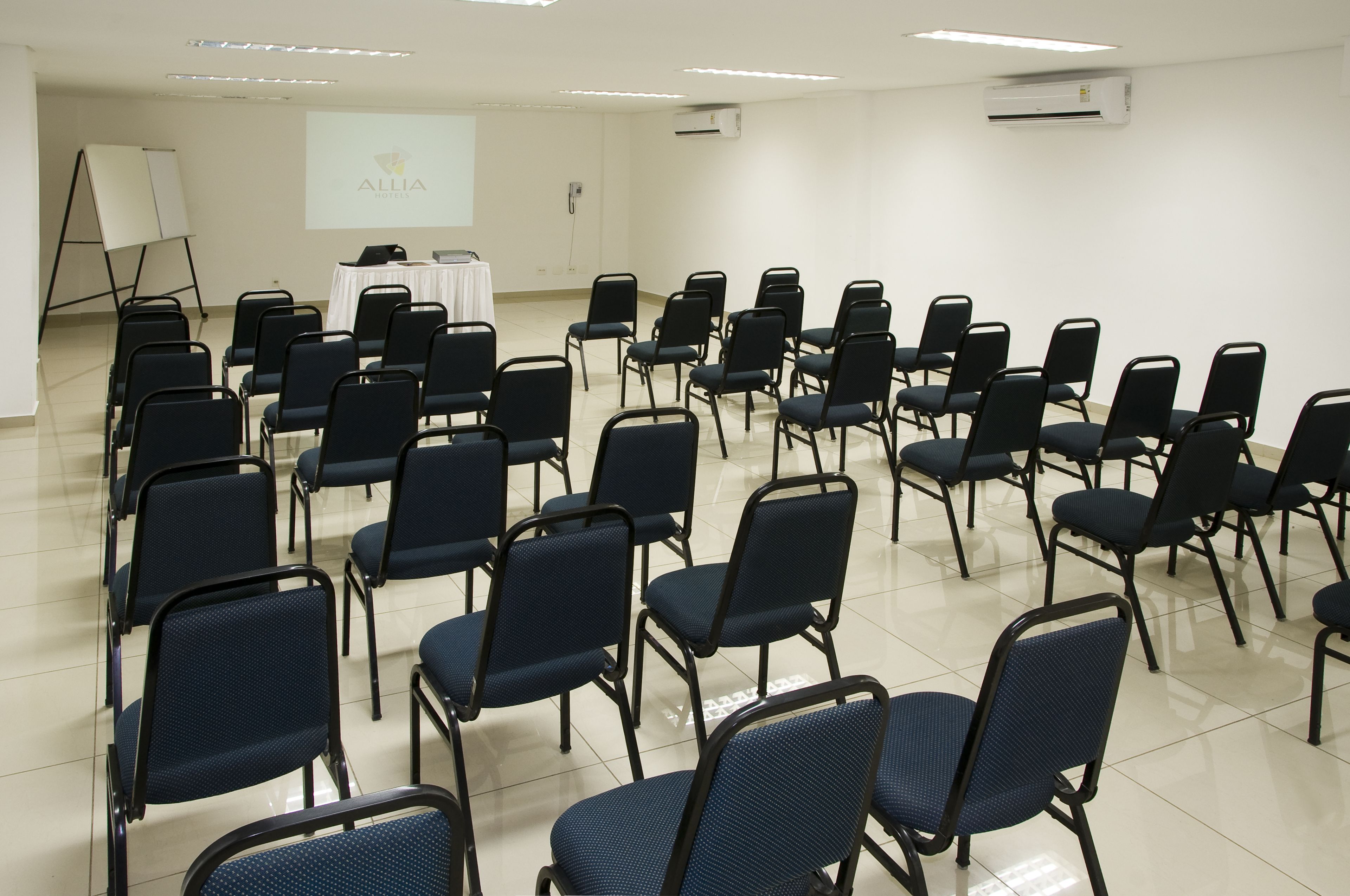 Uma sala de conferências com fileiras de cadeiras pretas, um quadro branco e uma tela de projeção exibindo o logotipo "ALLIA".