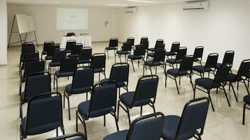 Uma sala de conferências com fileiras de cadeiras pretas, um quadro branco e uma tela de projeção exibindo o logotipo "ALLIA".