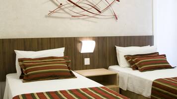 Quarto de hotel com duas camas, cada uma com roupa de cama e almofadas listradas, cabeceira de madeira, ar-condicionado fixado na parede e luminária simples.
