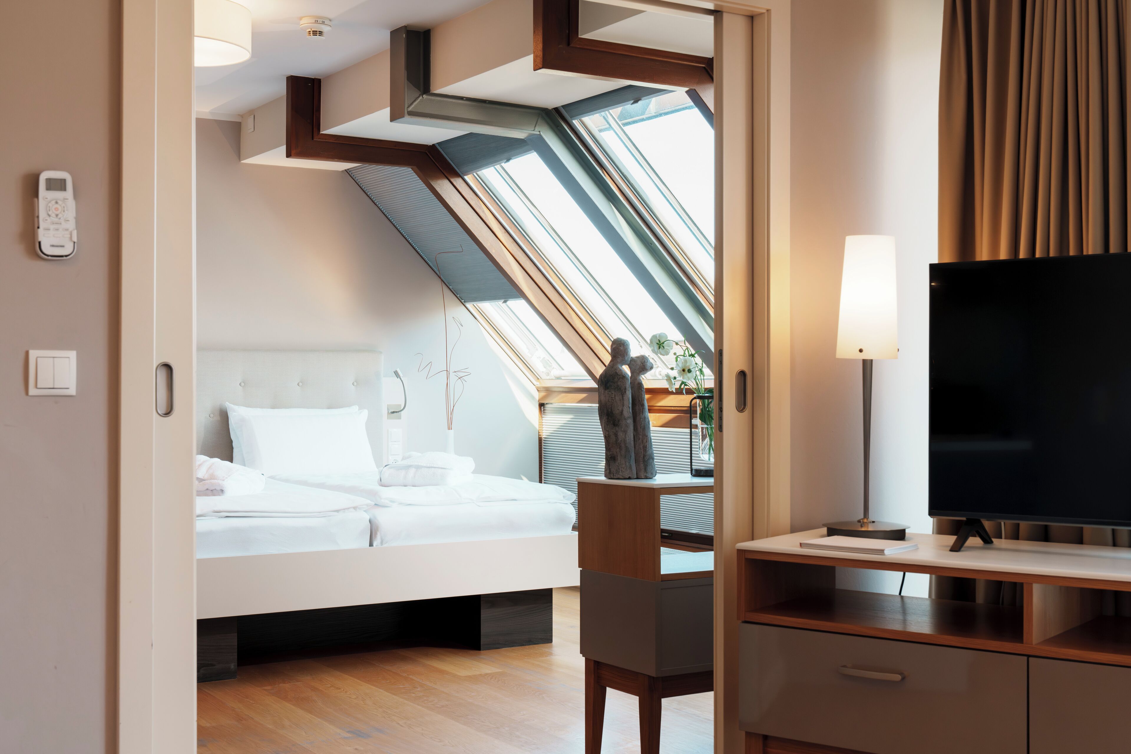 Quarto de hotel moderno com cama, escrivaninha e televisão, e claraboia.