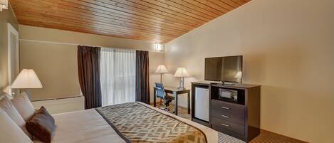 Quarto de hotel com teto de madeira, cama com roupa de cama estampada, escrivaninha com cadeira, televisão e minibar.